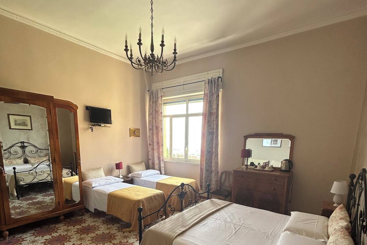 Quarto de hotel com uma cama grande, duas camas menores, um lustre, uma televisão e um espelho amplo.