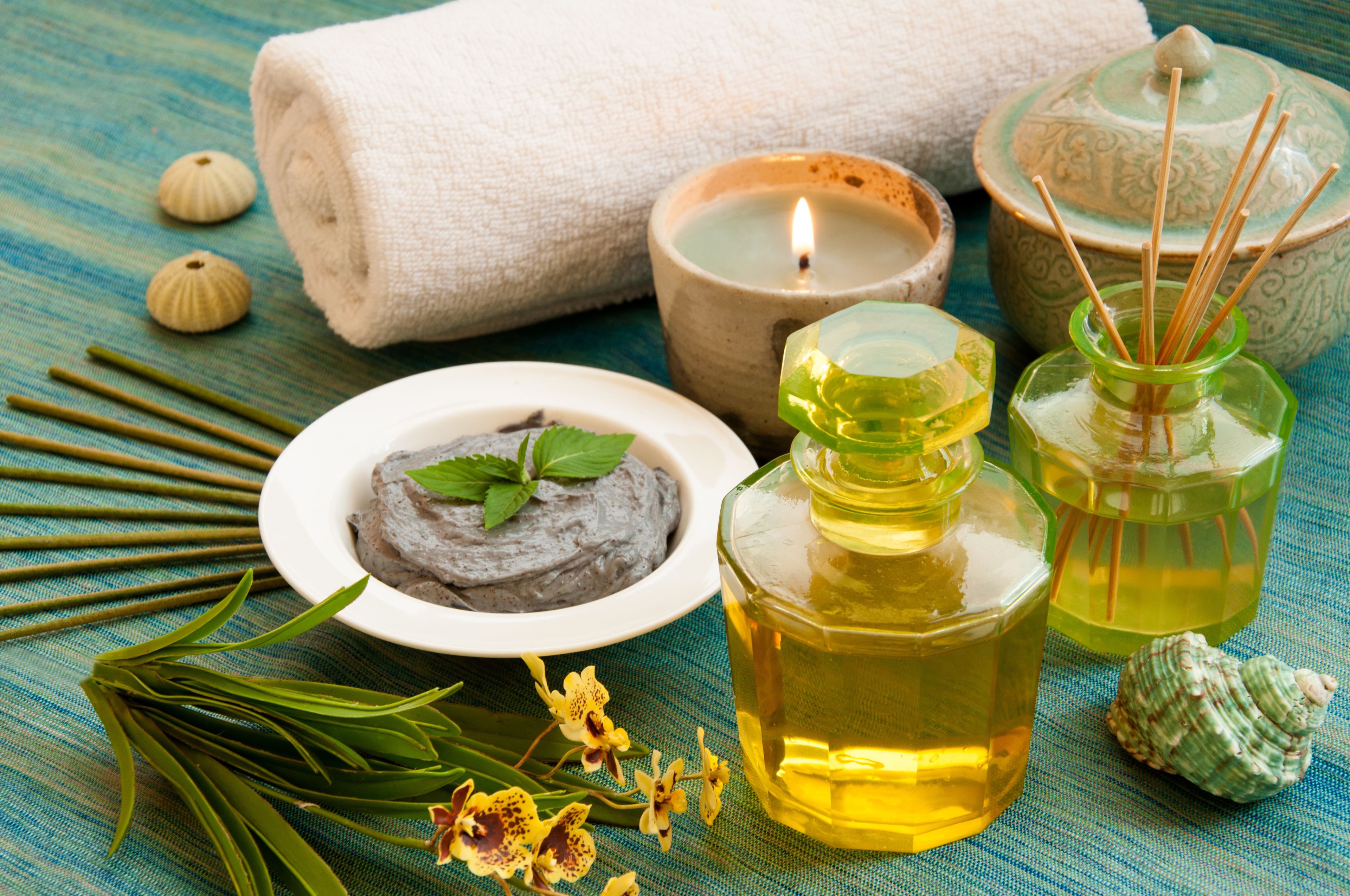 Ambiente de spa com uma tigela de máscara de argila preta, velas, frascos de óleo essencial e uma toalha enrolada sobre uma superfície azul texturizada.