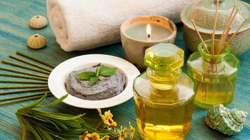 Ambiente de spa com uma tigela de máscara de argila preta, velas, frascos de óleo essencial e uma toalha enrolada sobre uma superfície azul texturizada.