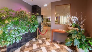Um saguão de hotel com piso xadrez, uma barreira de corda vermelha e plantas em vasos.