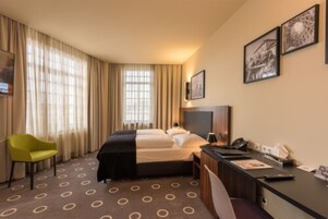 Quarto de hotel com cama, secretária, cadeira e janela com cortinas.