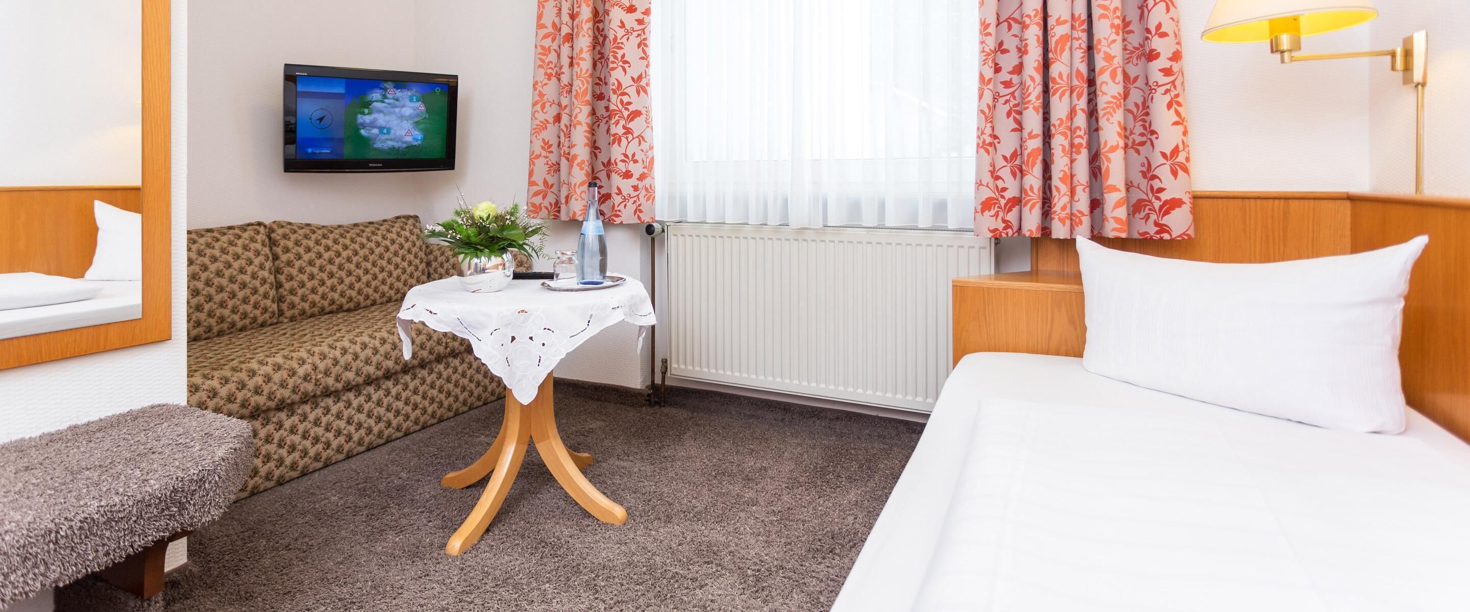 Quarto de hotel com uma cama, um sofá, uma mesa pequena com um vaso e uma garrafa, uma televisão na parede e cortinas.
