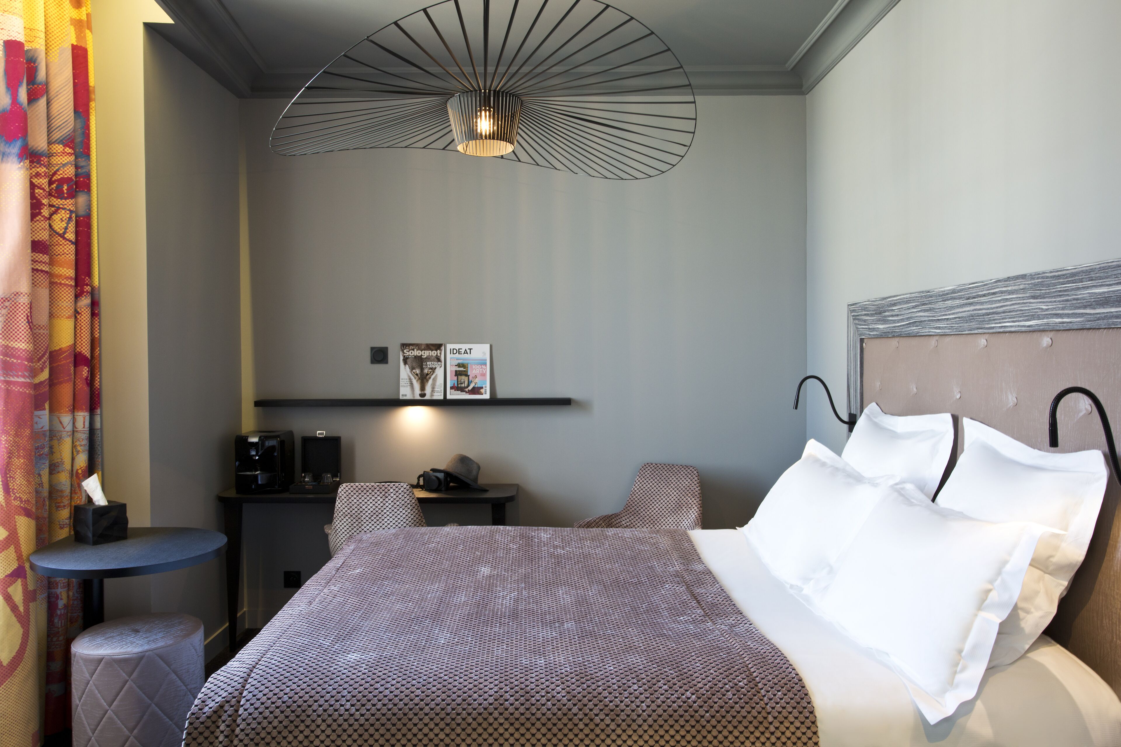 Quarto de hotel com uma cama, uma escrivaninha com cafeteira, uma cadeira, uma mesinha e uma prateleira fixada na parede com moldura para foto.