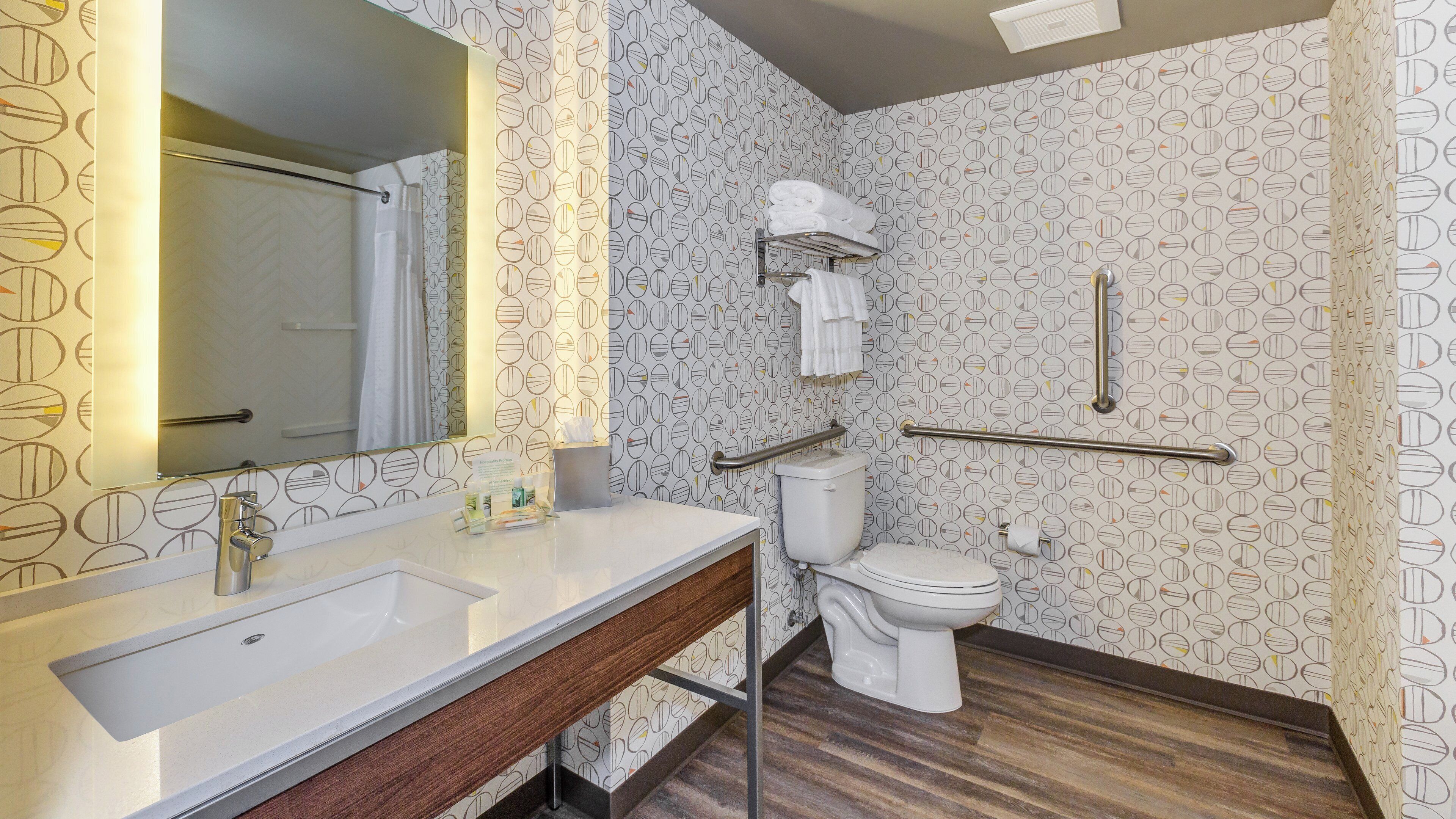 Casa de banho com lavatório branco, espelho e sanita. Inclui barras de apoio e um porta-toalhas.