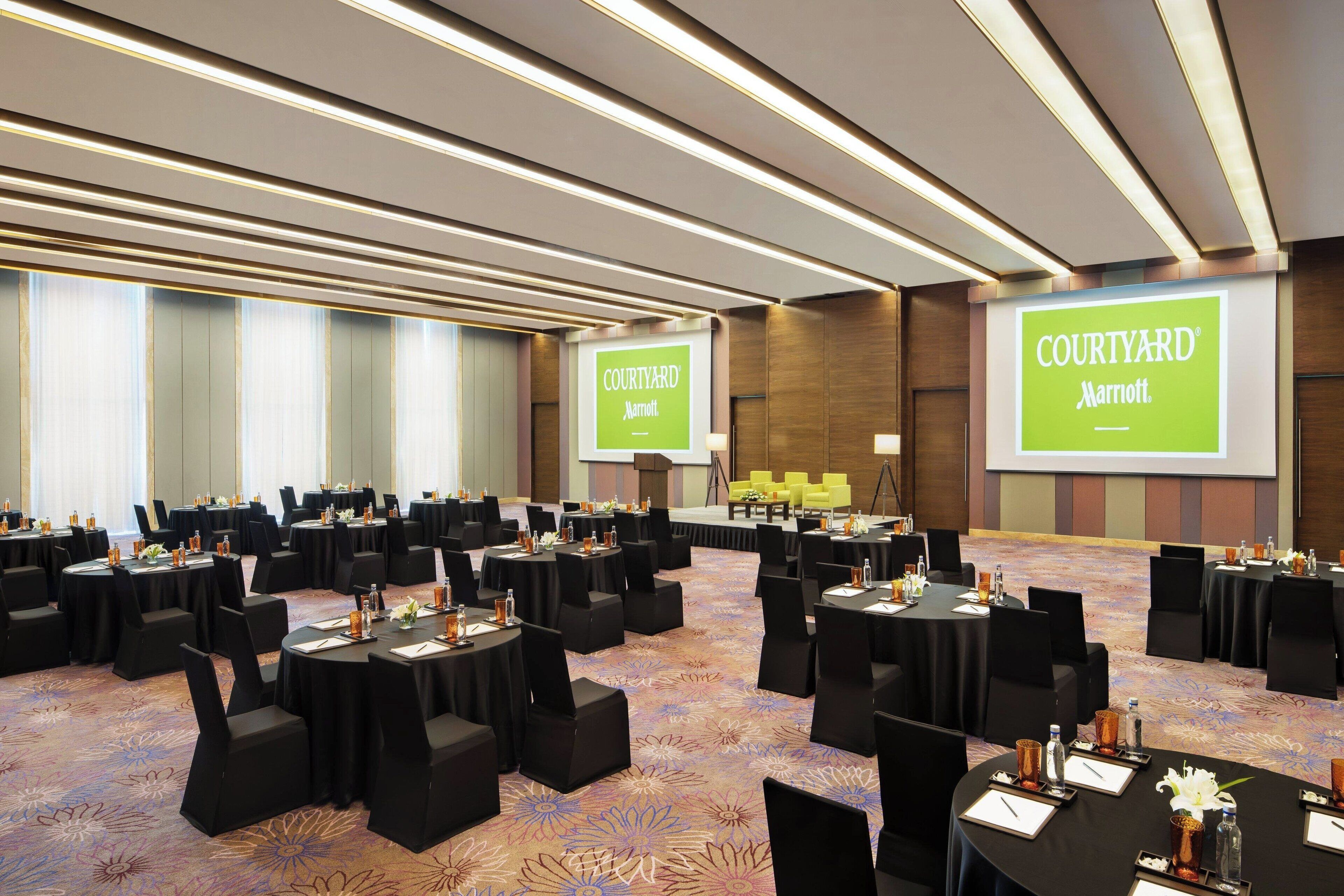 Uma sala de conferências com várias mesas e cadeiras, uma tela grande exibindo 'Courtyard Marriott' e um palco com um púlpito.