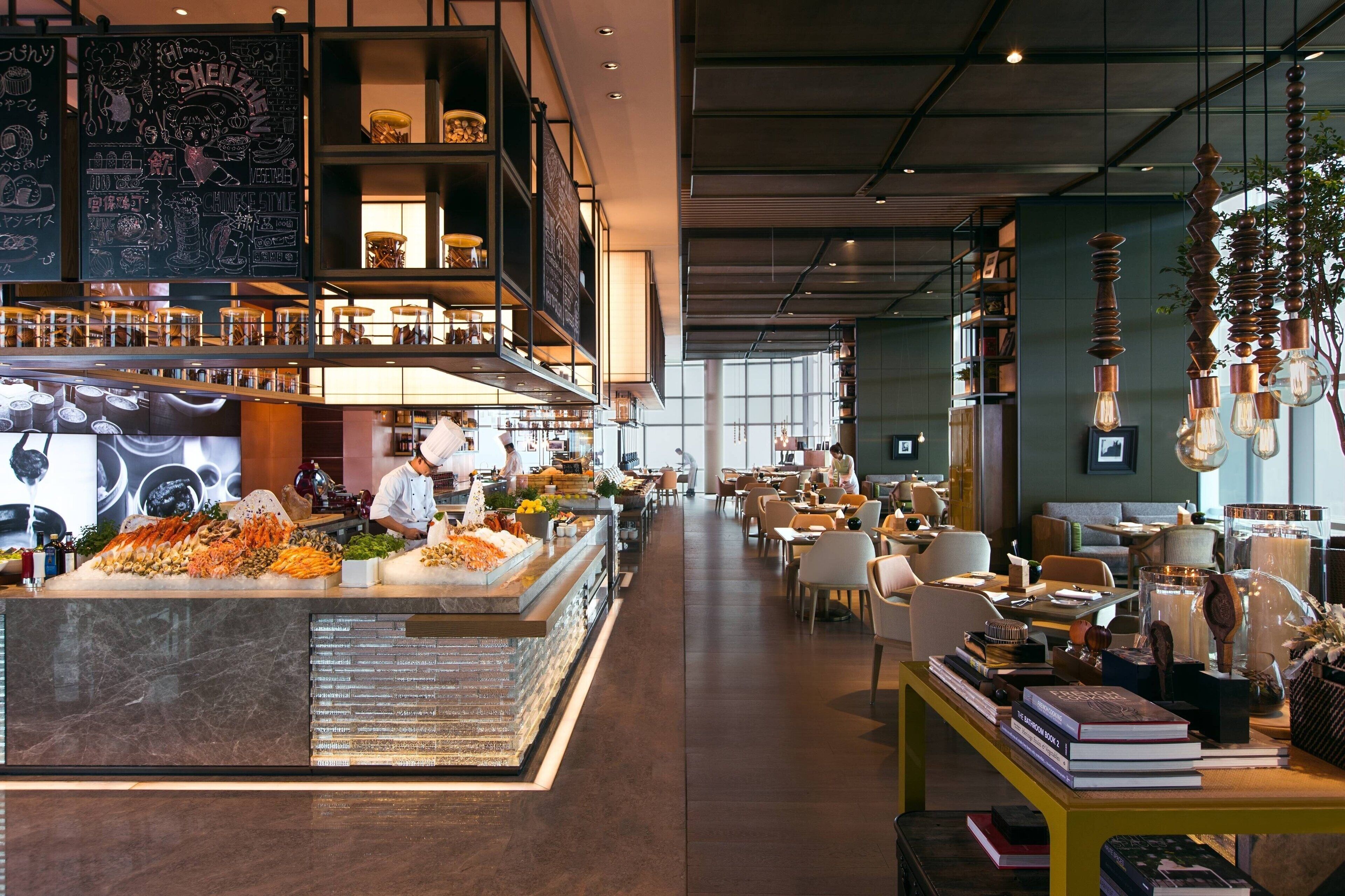 Um restaurante moderno com um balcão central exibindo alimentos, área de bar e sala de estar com mesas e cadeiras.