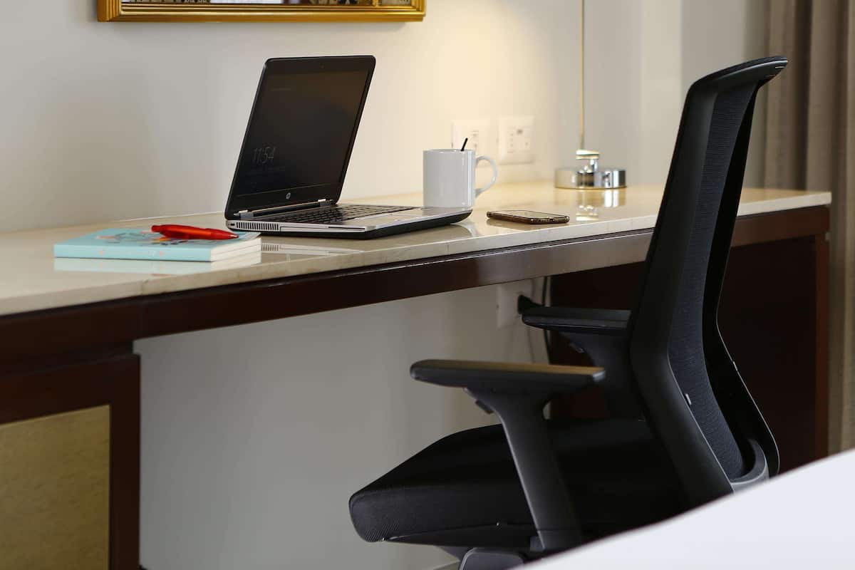 Um espaço de trabalho em hotel com um laptop, uma caneca branca, um smartphone e uma cadeira.