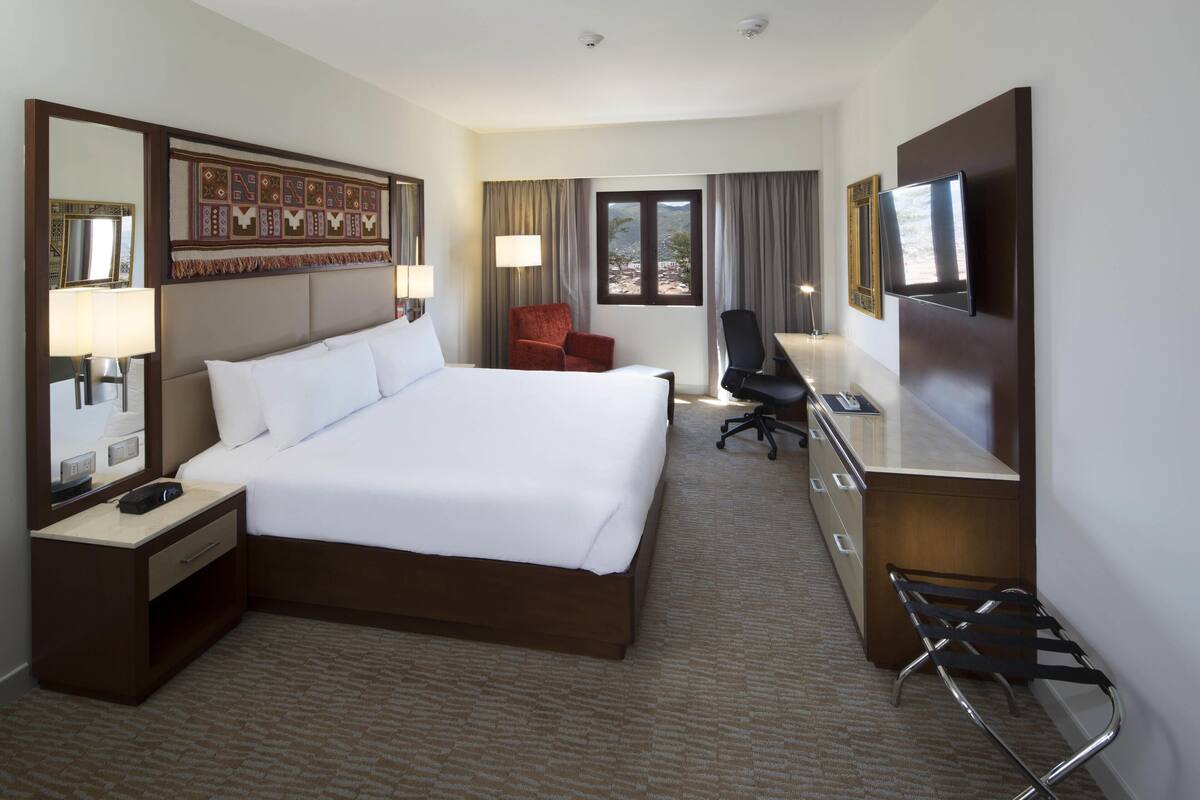 Quarto de hotel com uma cama grande, uma escrivaninha com cadeira, televisão e janela com cortinas.