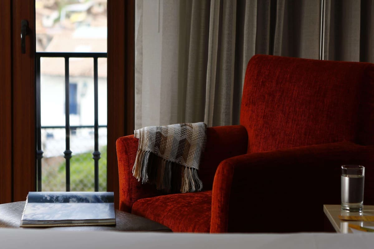 Quarto de hotel com uma poltrona vermelha, vista de edifícios pela janela, uma luminária e um copo de água sobre uma mesa.