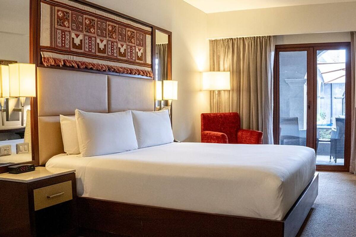 Quarto de hotel com uma cama grande, uma cadeira vermelha, uma mesa e vista para o exterior através de uma porta de vidro.
