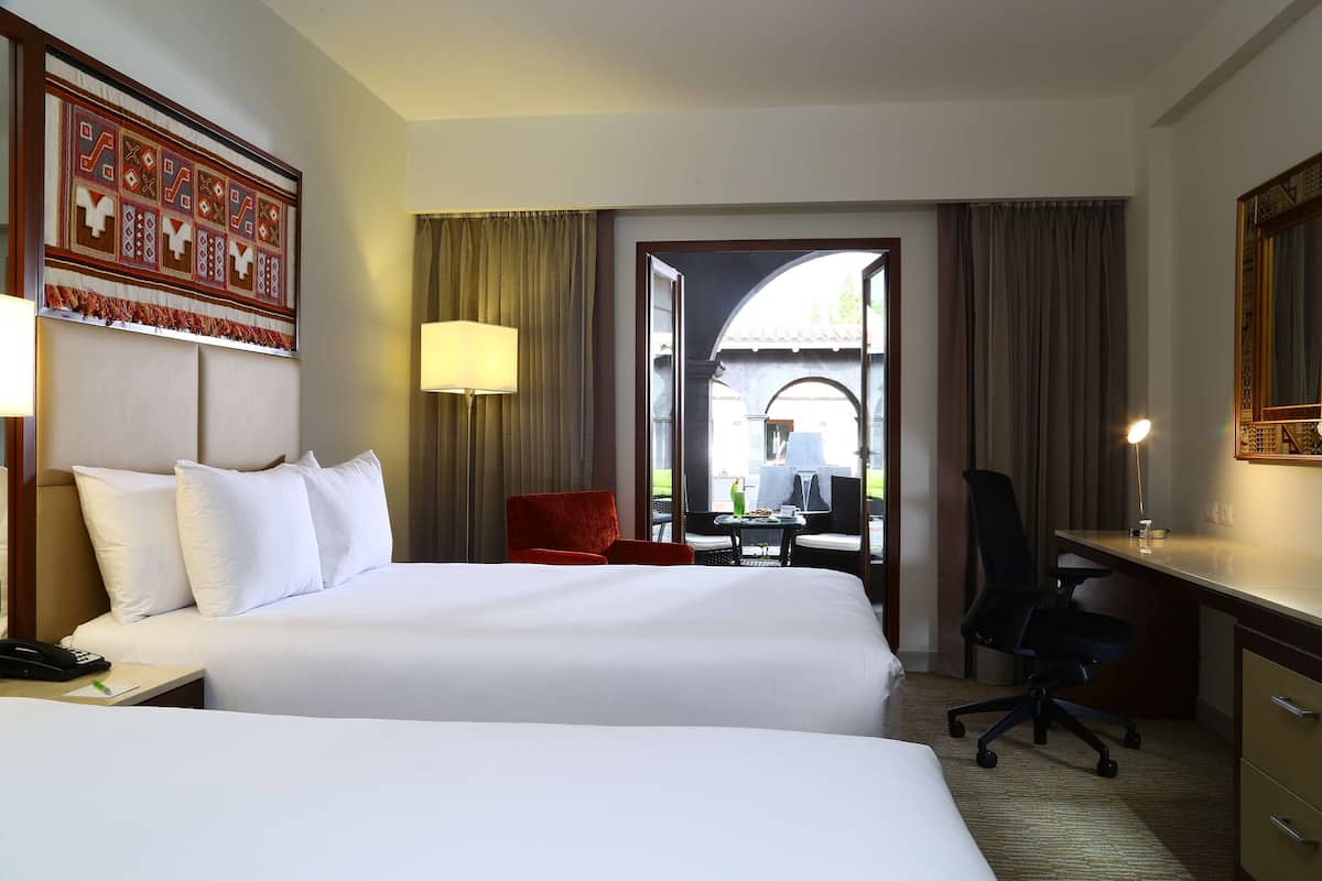 Quarto de hotel com duas camas, uma escrivaninha, uma cadeira e vista para um pátio externo.