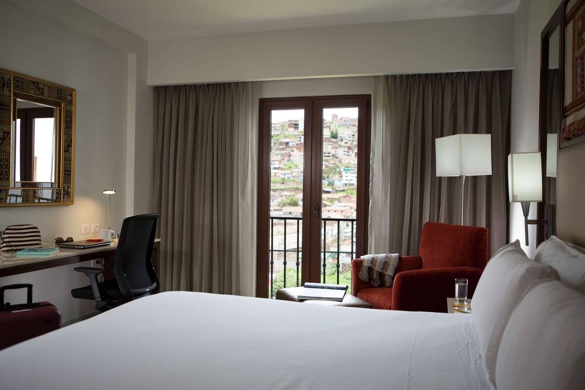 Quarto de hotel com uma cama grande, uma escrivaninha com cadeira, uma poltrona vermelha, uma mesinha de centro com um copo de água e vista para a cidade pela janela.