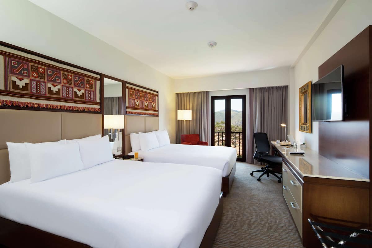 Quarto de hotel com duas camas, uma escrivaninha, uma cadeira, televisão e varanda com vista.