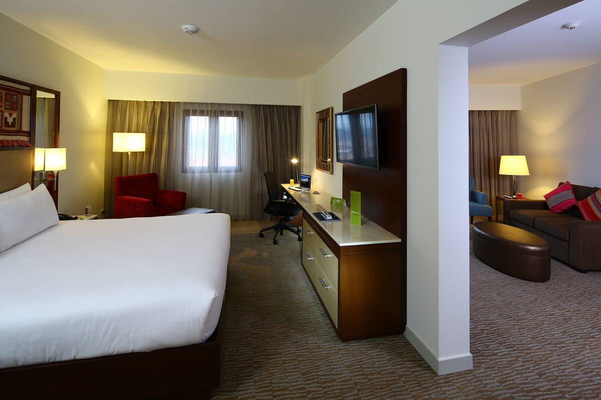Quarto de hotel com uma cama grande, uma escrivaninha com computador, televisão e um espaço de estar com sofá e poltrona.