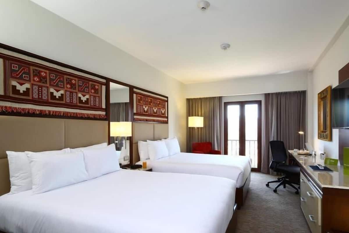 Quarto de hotel com duas camas, uma escrivaninha, uma cadeira, uma televisão e uma janela grande.