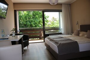 Quarto de hotel com uma cama, uma escrivaninha, uma cadeira, varanda com vista para árvores e janela com cortinas.