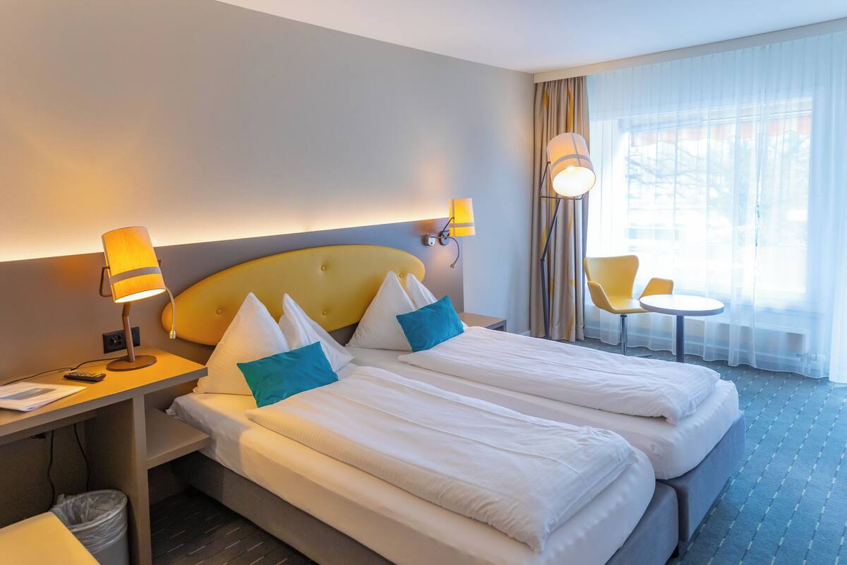 Quarto de hotel com duas camas, uma escrivaninha com abajur, uma cadeira e uma mesinha.