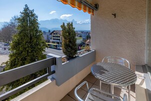 Varanda com mesa e cadeiras de metal, com vista para uma rua com edifícios e montanhas ao fundo.