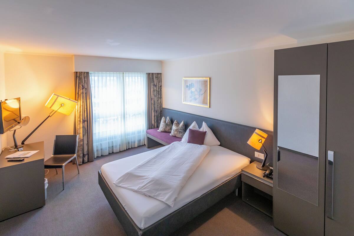 Quarto de hotel com cama, mesa de cabeceira, escrivaninha, cadeira e guarda-roupa.