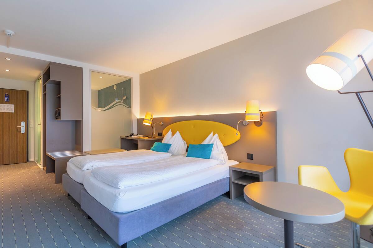Quarto de hotel com cama, mesas de cabeceira, escrivaninha, cadeira e abajur.
