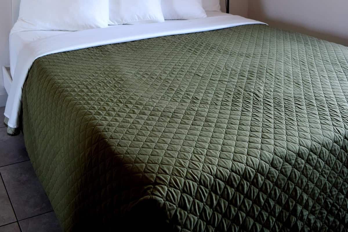 Cama bem arrumada com roupa de cama branca e manta acolchoada verde, luminária de cabeceira e quadros nas paredes.