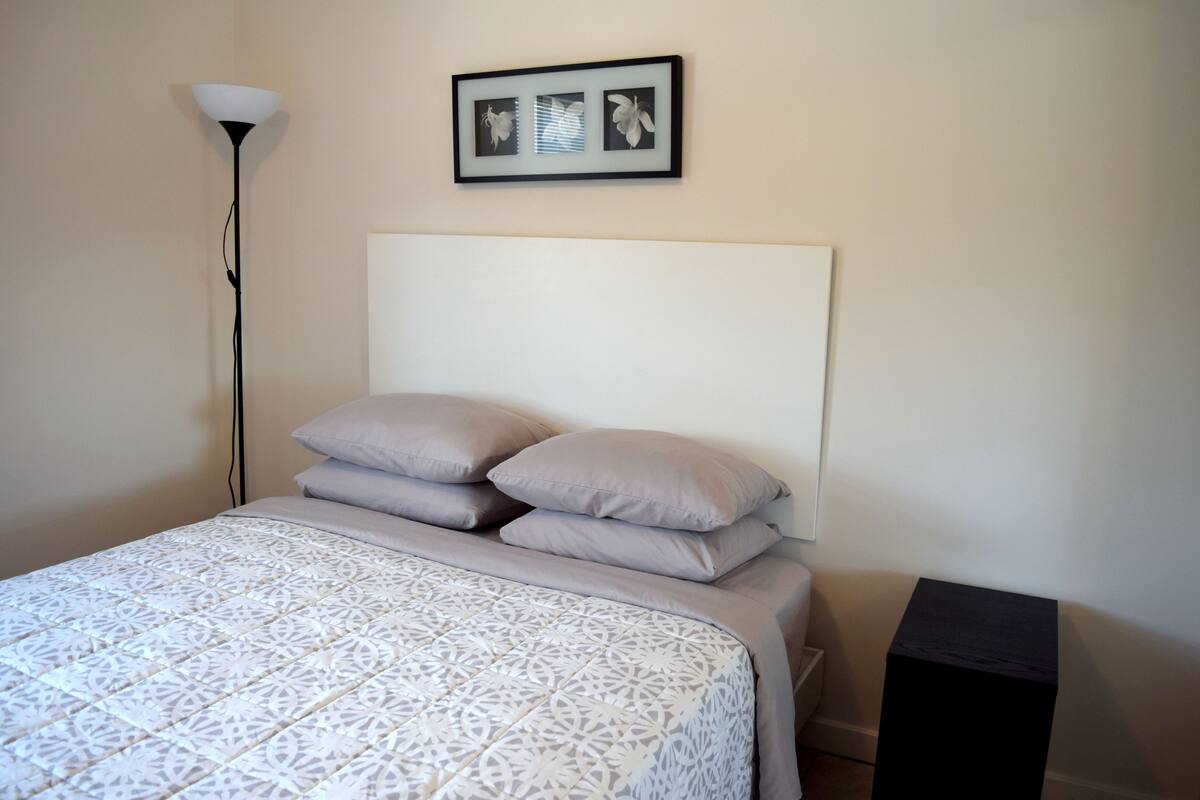 Quarto com cama, almofadas, um quadro na parede e uma luminária de chão.