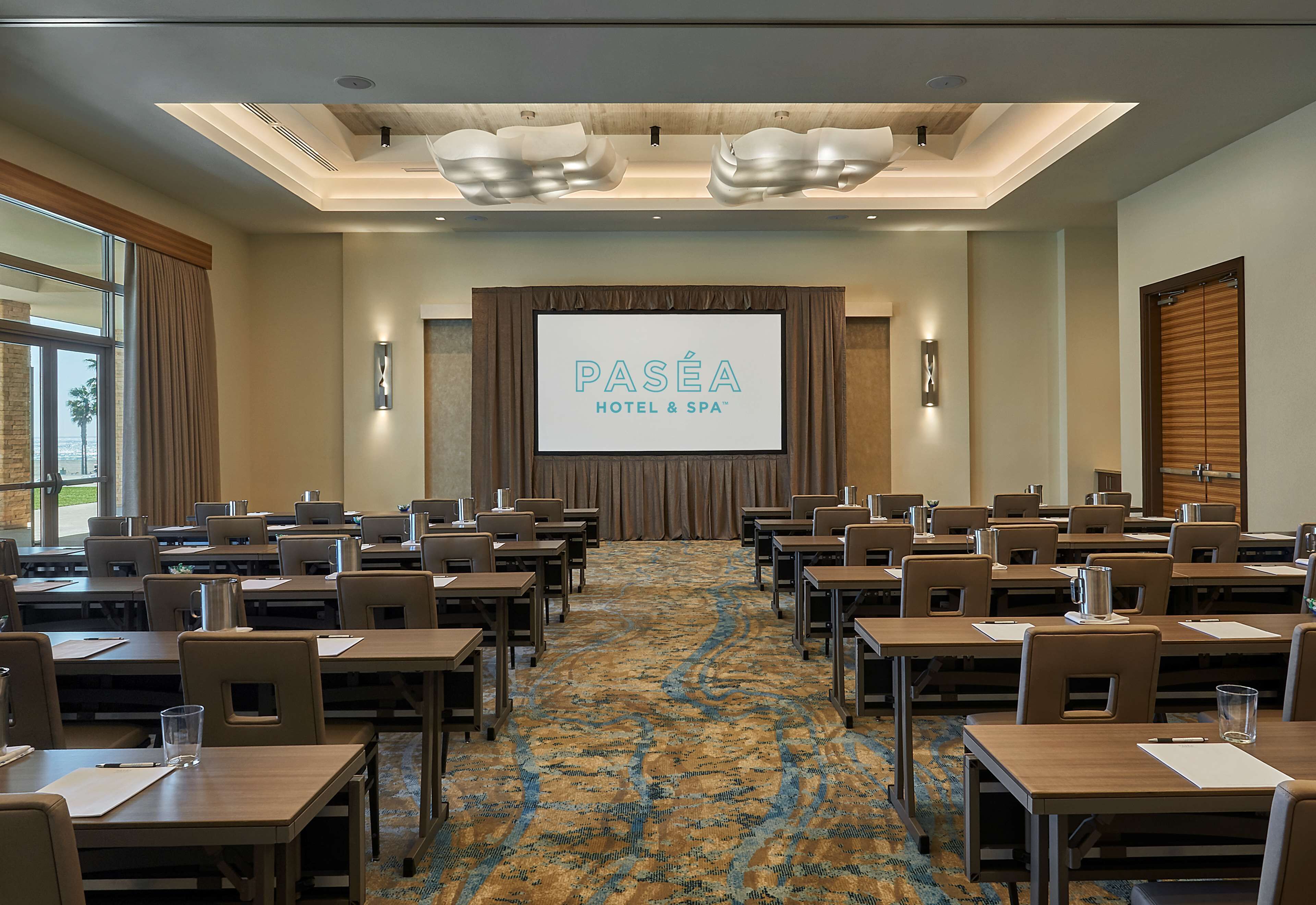 Uma sala de conferências com uma tela grande exibindo 'PASÉA HOTEL & SPA' na frente. Fileiras de mesas e cadeiras organizadas para uma reunião ou apresentação.