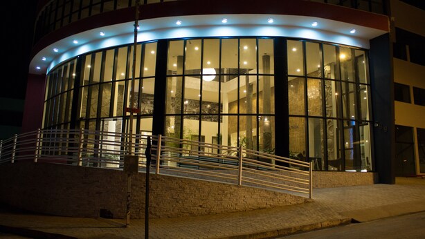 Um edifício moderno com fachada de vidro curva, uma escadaria que leva à entrada e exterior bem iluminado.