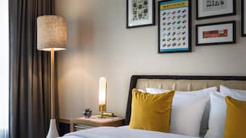 Quarto de hotel com uma cama, uma mesa de cabeceira com abajur, uma coleção de caixa de fósforos emoldurada e uma janela grande com cortinas.
