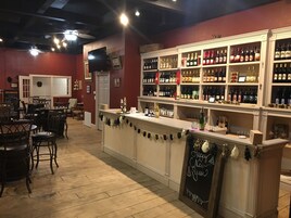 Uma área de bar com cadeiras e mesas de madeira, um balcão com um letreiro de 'Feliz Ano Novo' e prateleiras cheias de garrafas de vinho.