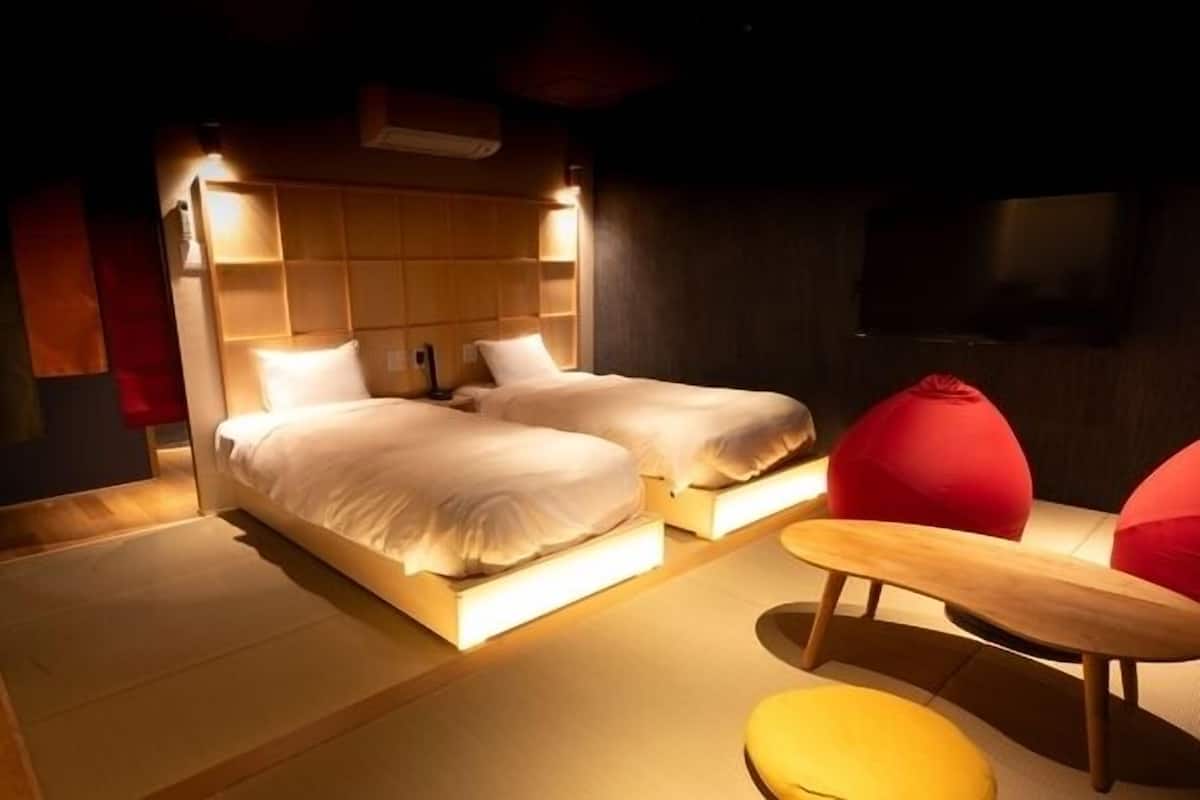 Quarto de hotel moderno com duas camas, uma mesinha e uma cadeira vermelha.