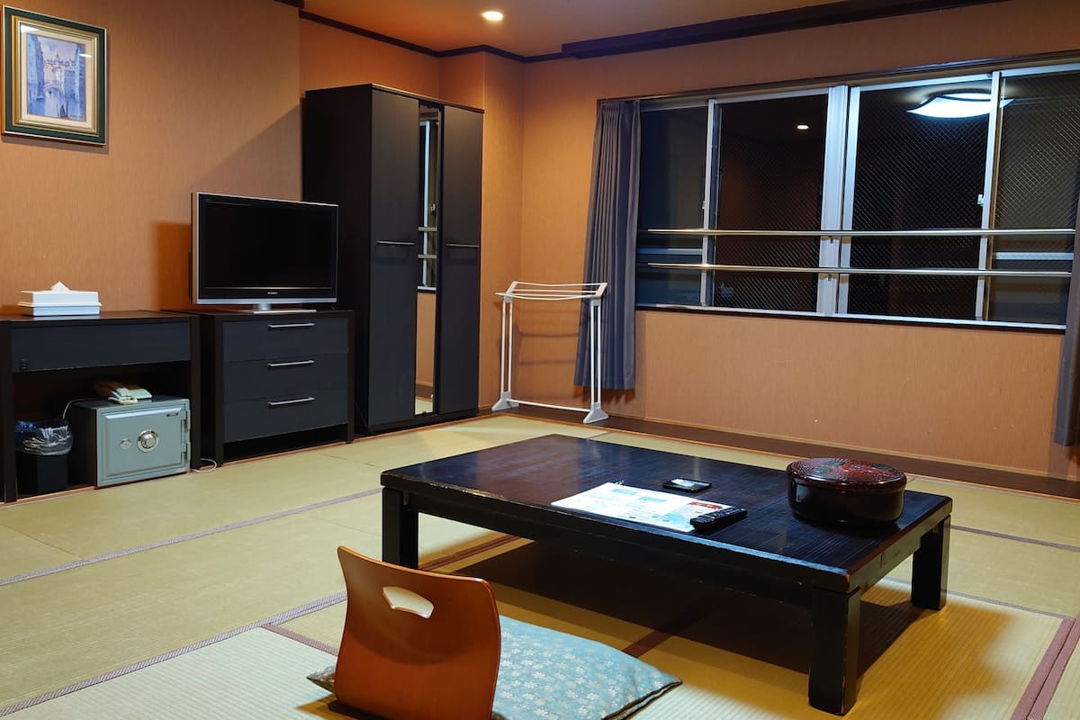 Quarto com piso de tatame, um suporte de TV preto, um armário, uma janela com cortinas e uma mesa tradicional japonesa.