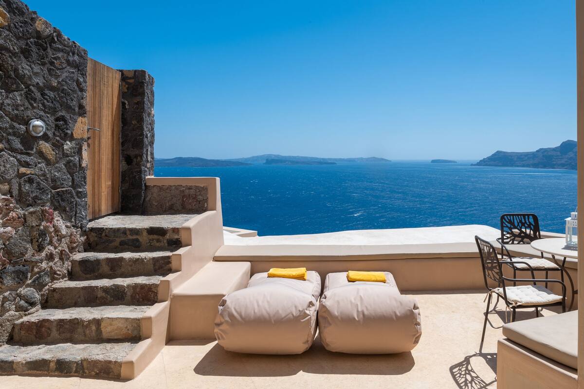 Escadaria de pedra que leva a um terraço com duas áreas de estar acolchoadas e uma mesa, com vista para um mar azul e límpido.