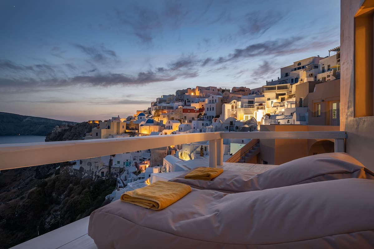 Varanda com cama e toalhas, com vista para uma vila costeira de edifícios brancos e mar.