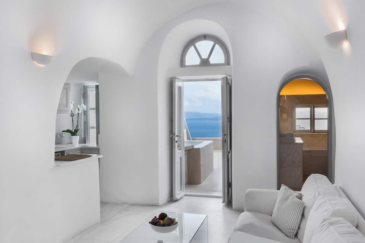 Sala de estar moderna e minimalista com um sofá branco, uma mesa de centro com frutas e vista para o mar através de uma porta aberta.