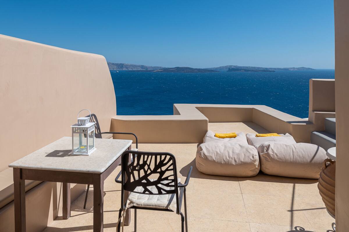 Varanda com mesa, cadeiras e almofadas, com vista para um corpo de água e terra distante.