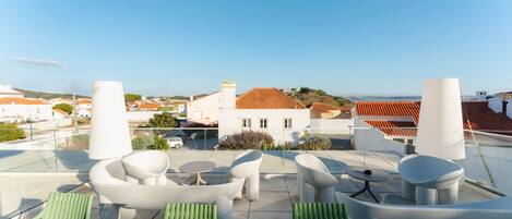 Terraço no telhado com mobiliário branco moderno, vista ampla da paisagem urbana e um edifício tradicional com telhado vermelho ao fundo.