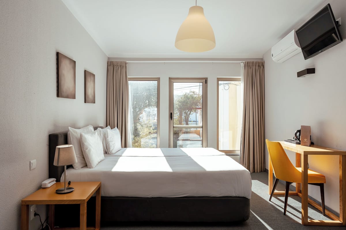 Quarto de hotel com cama, escrivaninha, cadeira, luminária, televisão e ar condicionado.