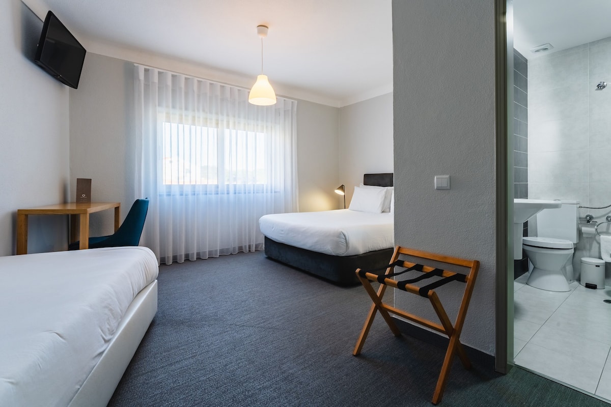 Quarto de hotel com duas camas, uma escrivaninha, uma cadeira, televisão, janela com cortinas e banheiro com vaso sanitário e pia.