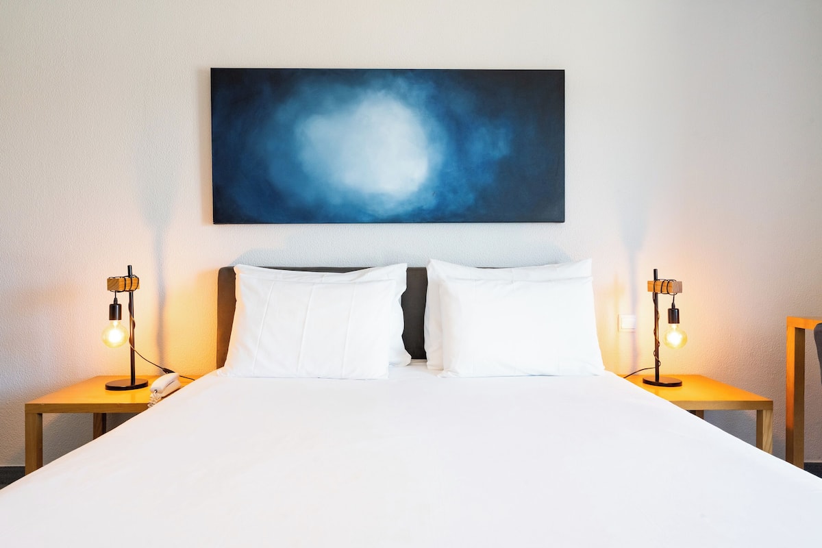 Uma cama bem arrumada com lençóis brancos, acompanhada por duas mesas de cabeceira, cada uma com um abajur diferente. Um grande quadro abstrato enfeita a parede acima da cama.