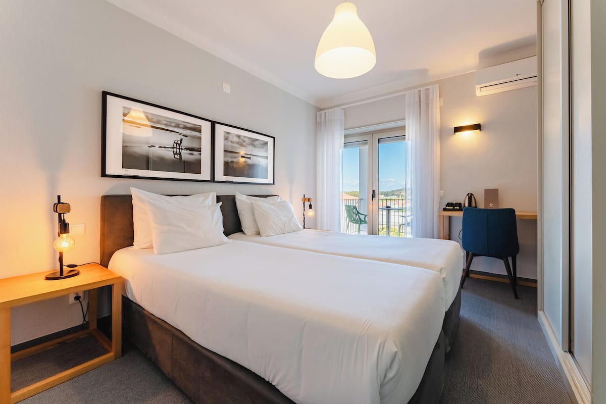 Quarto de hotel com duas camas, uma mesa com cadeira, varanda com vista e quadros nas paredes.