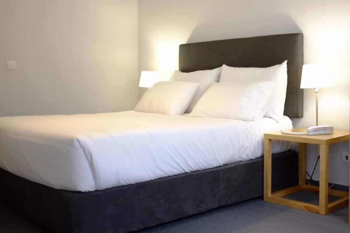 Cama bem arrumada com lençóis brancos e cabeceira escura, acompanhada por duas luminárias de mesa em uma mesa de cabeceira de madeira.