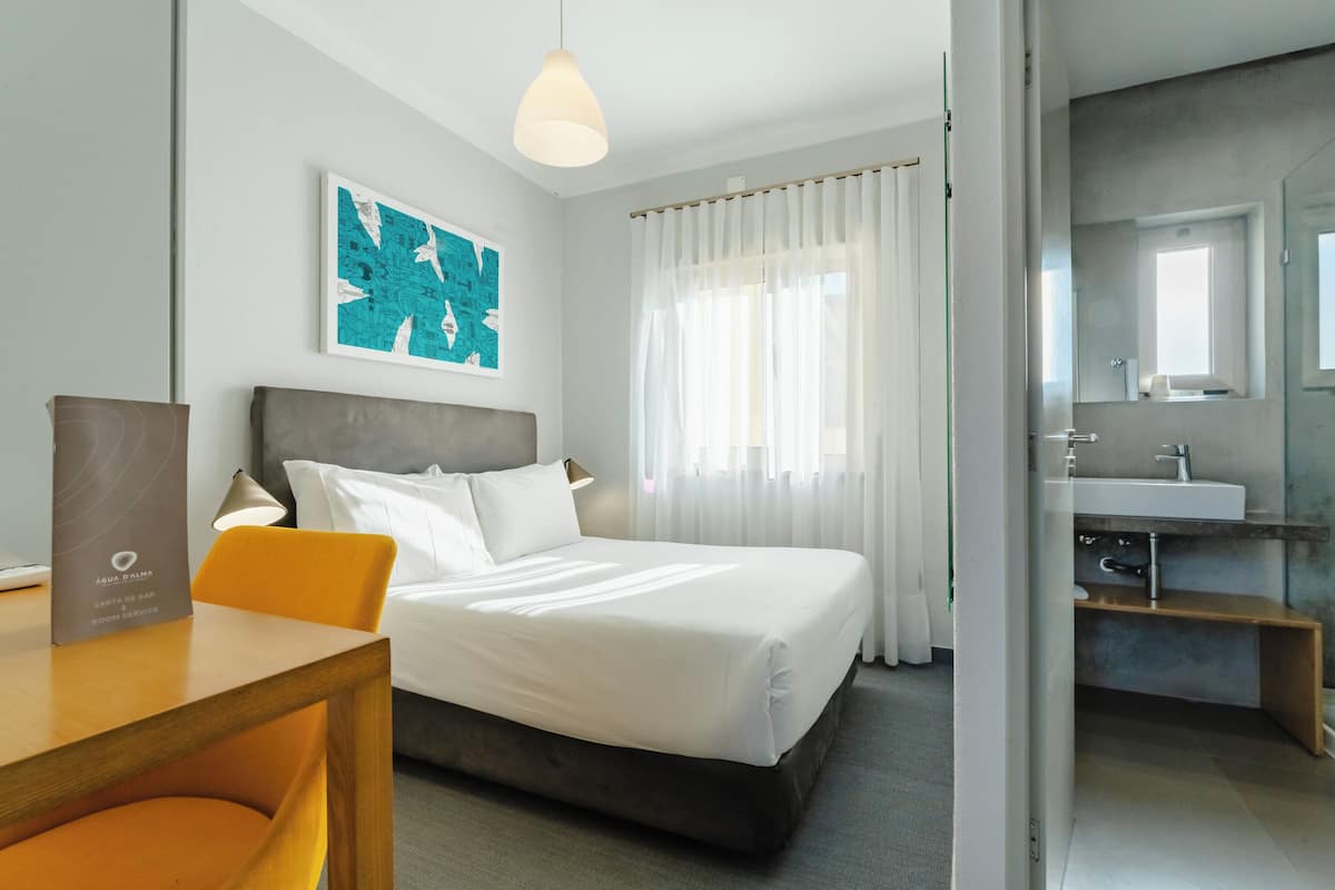 Quarto de hotel com cama, escrivaninha, cadeira, mesa de cabeceira, luminária, janela com cortinas, banheiro com pia e espelho, e uma obra de arte na parede.