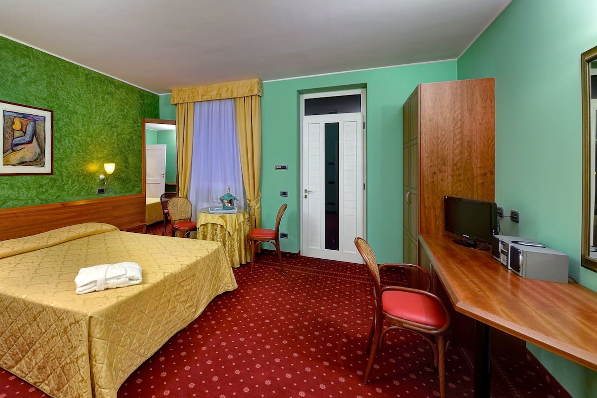 Quarto de hotel com cama, escrivaninha, cadeira, mesinha de cabeceira com abajur e televisão.