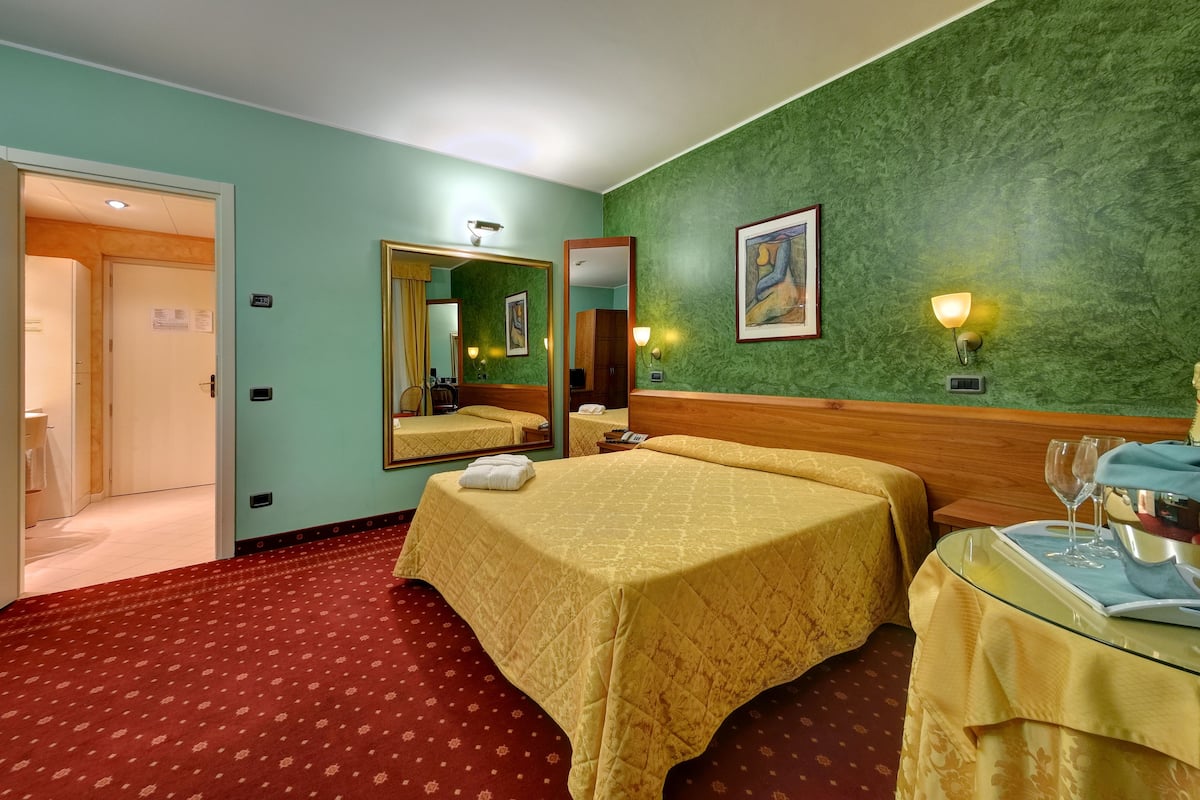 Quarto de hotel com duas camas, um espelho, um quadro e uma mesa com uma garrafa e copos.