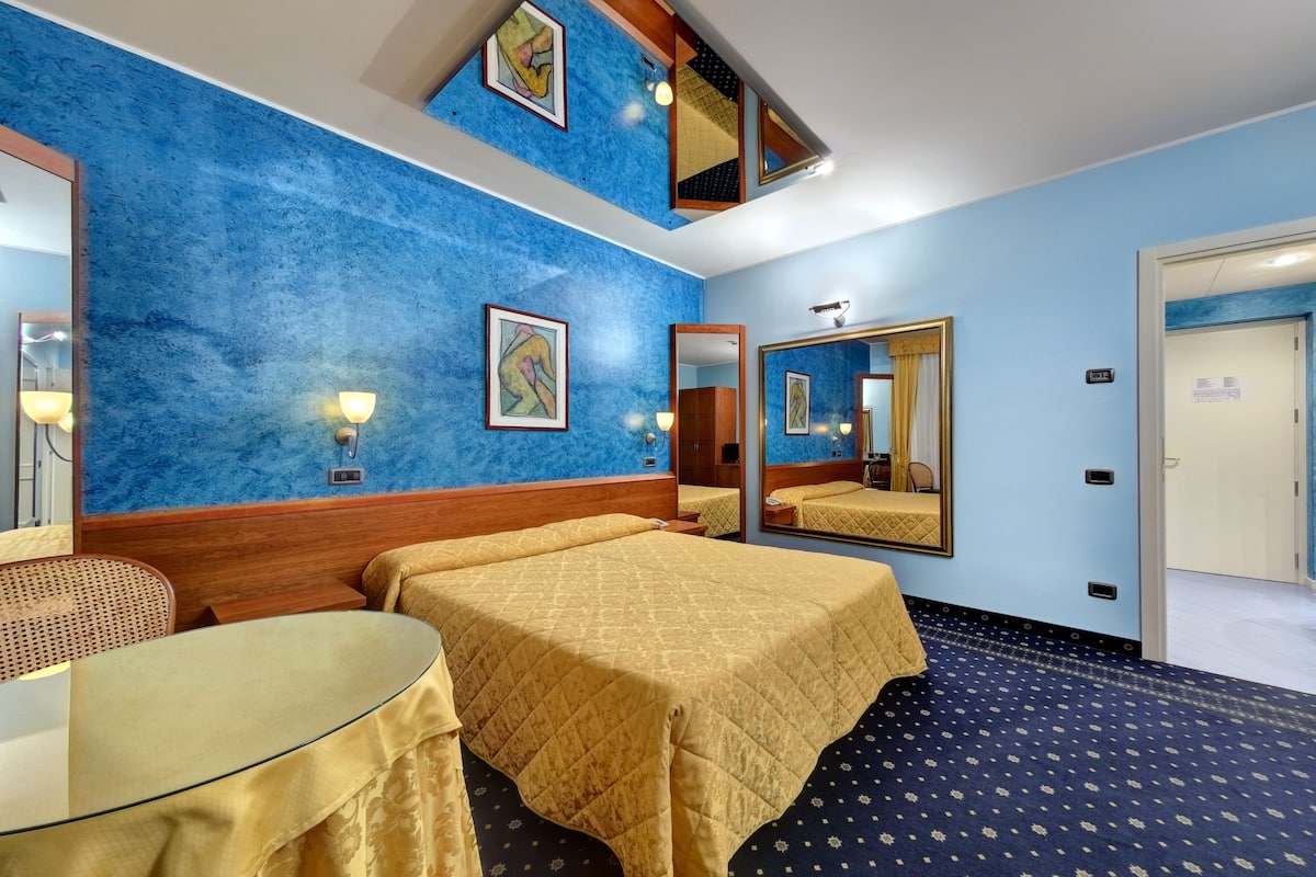 Quarto de hotel com duas camas, uma mesa de vidro e tapete azul.