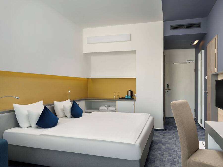 Quarto de hotel com cama, escrivaninha, cadeira, televisão e banheiro visível por uma porta aberta.