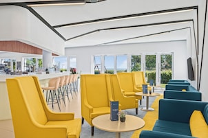 Uma área de estar moderna com assentos amarelo e azul, um bar com banquetas e grandes janelas com vista para o exterior.
