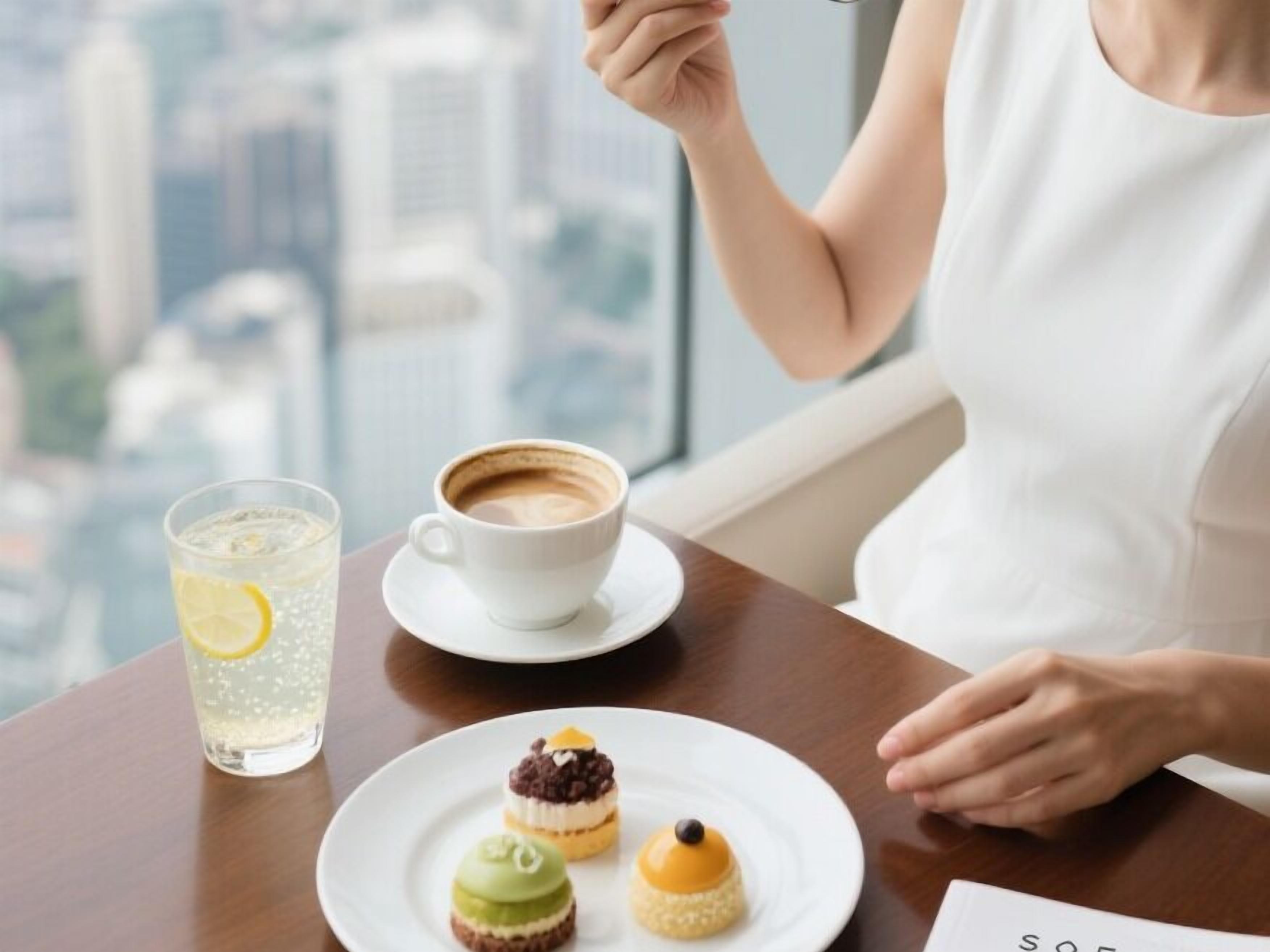 Uma pessoa sentada à mesa com uma xícara de café, um copo de chá gelado com uma fatia de limão e três pequenas sobremesas num prato.