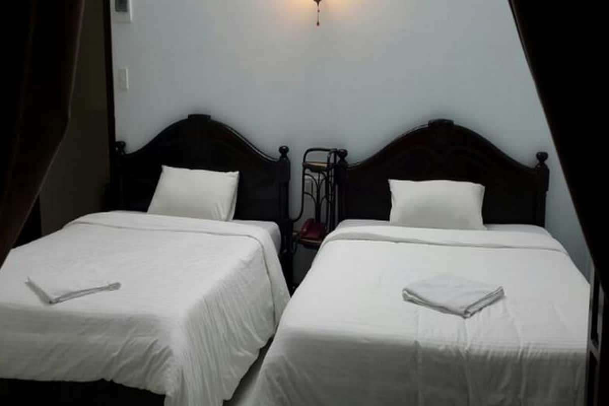 Duas camas de solteiro com roupa de cama branca, uma cabeceira de madeira escura e uma luminária fixa na parede.